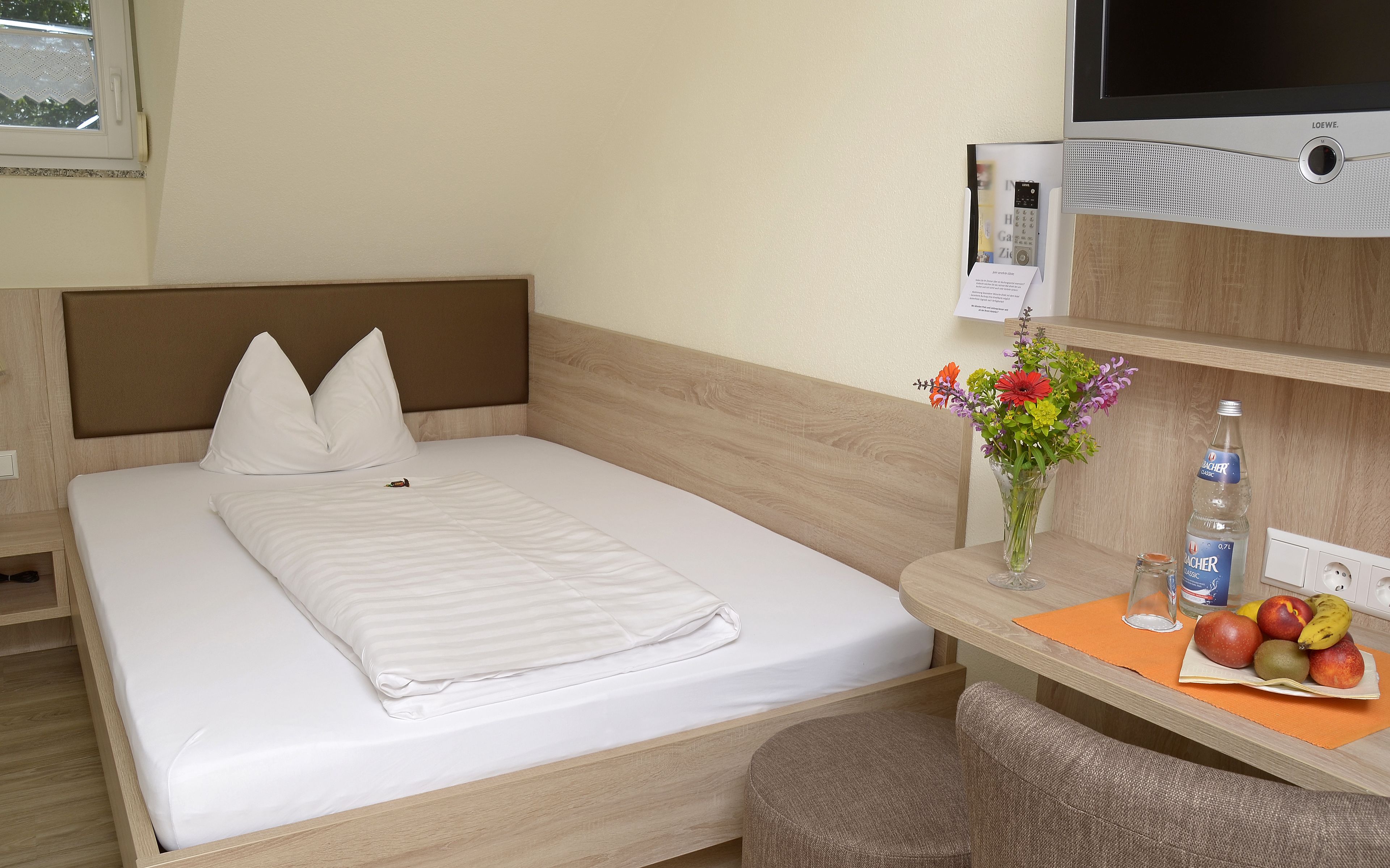 Cama bem arrumada com lençóis brancos, cabeceira de madeira, mesa de cabeceira pequena com um vaso de flores, uma garrafa de água e uma cesta de frutas.