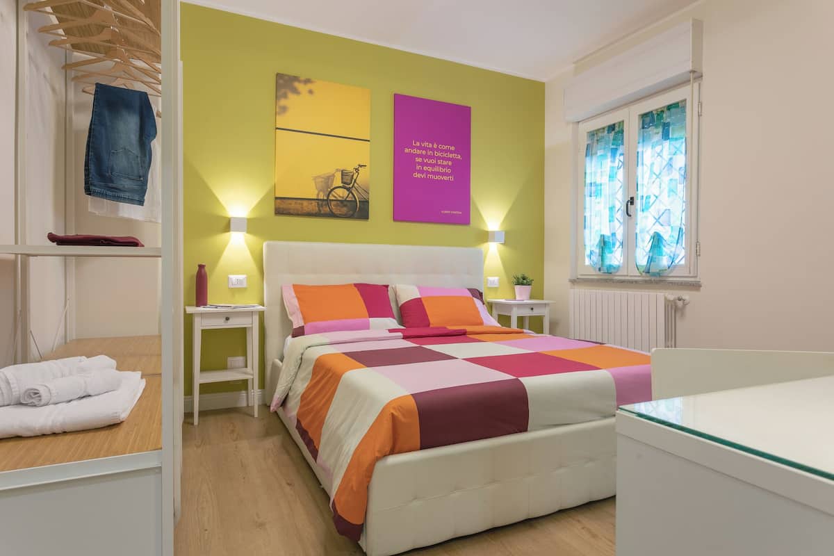 Quarto com cama, mesas de cabeceira, janela com cortinas, parede com um pôster e um cabideiro com roupas.