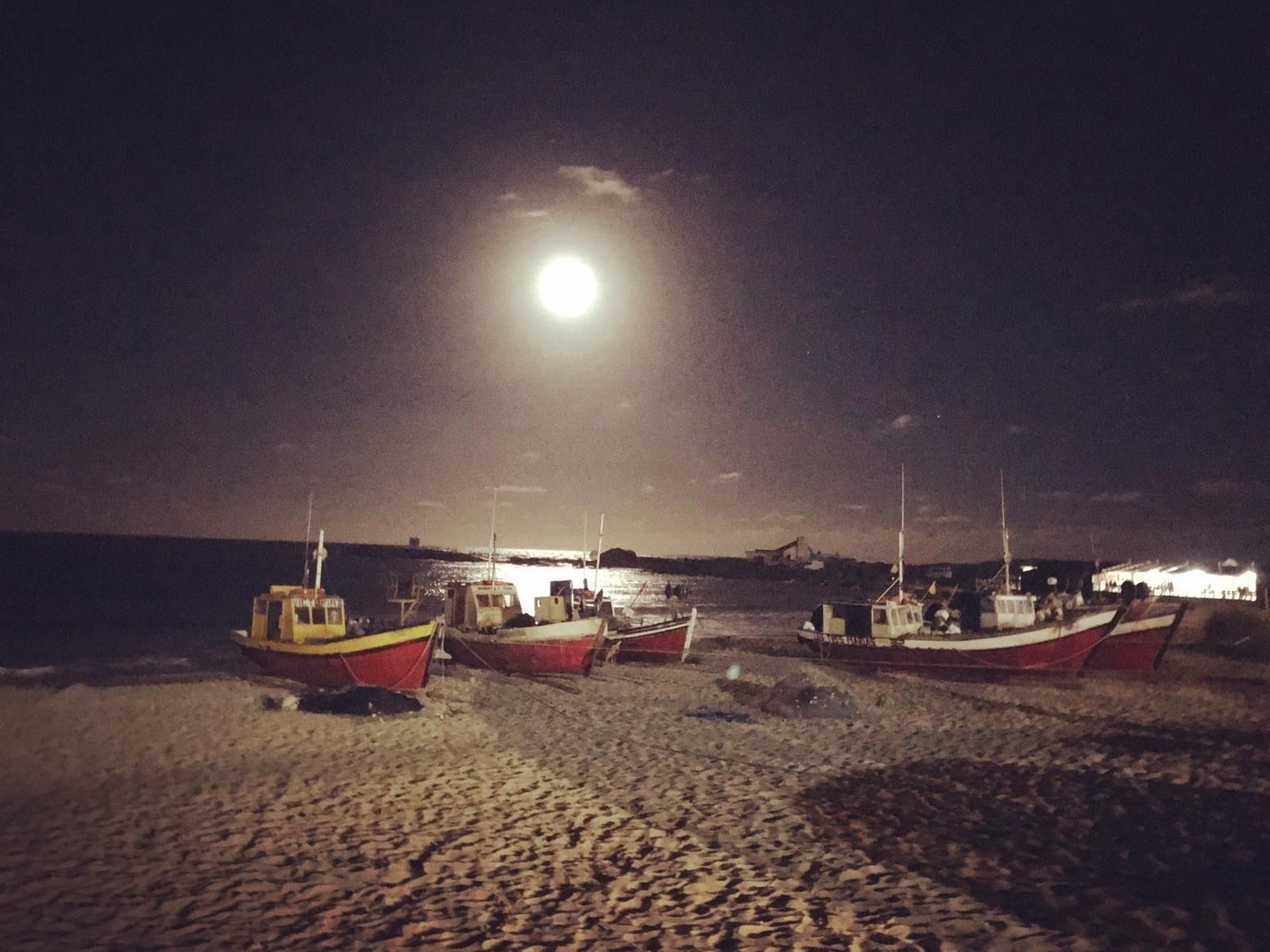 Barcos numa praia à noite com uma lua cheia no céu.