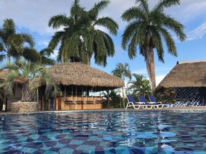 Uma piscina com espreguiçadeiras e estruturas de palha, cercada por palmeiras.