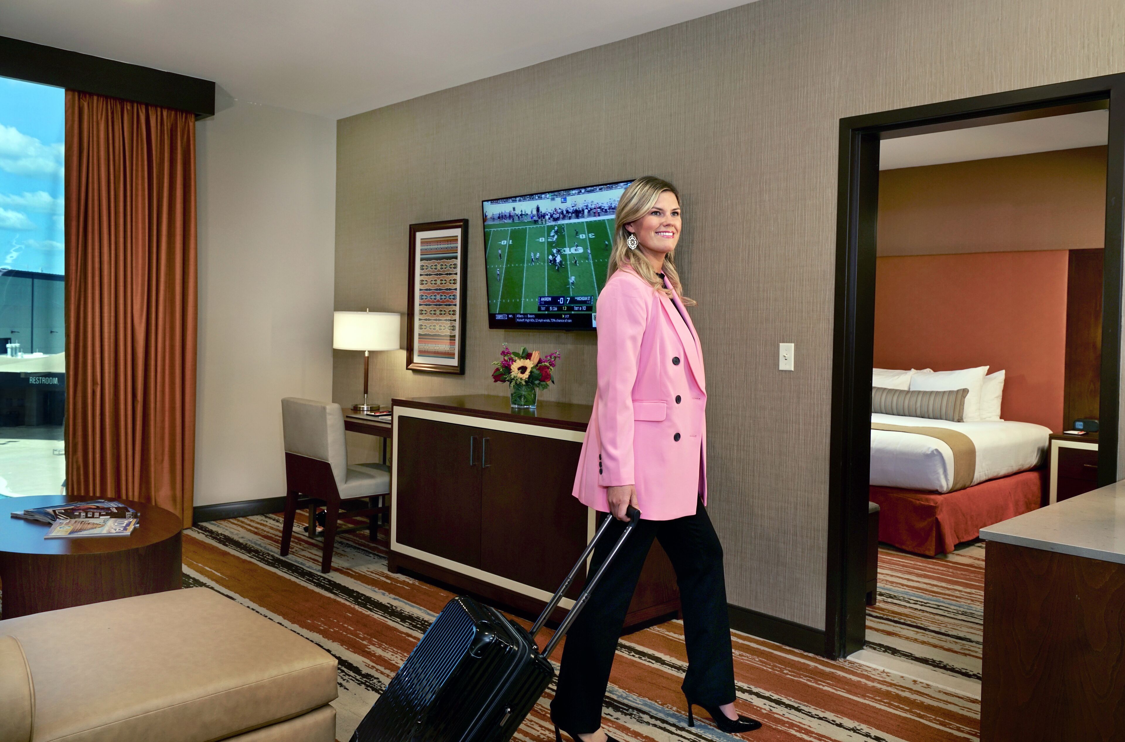 Quarto de hotel com cama, secretária com televisão, poltrona, sofá e janela com cortinas.