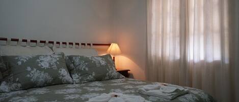 Cama bem arrumada com roupa de cama florida, abajur e janela com cortinas leves.