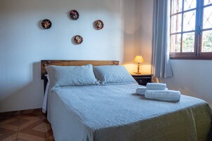 Uma cama bem arrumada com colcha e almofadas brancas, uma mesinha de cabeceira com abajur e três objetos decorativos fixados na parede.