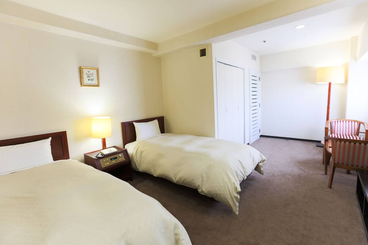 Quarto de hotel com duas camas, uma escrivaninha, uma cadeira, um abajur e uma porta.