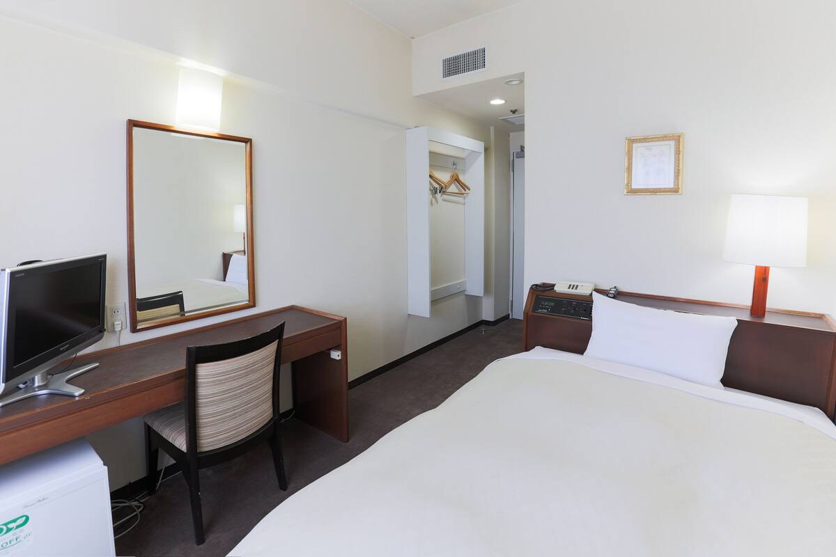 Quarto de hotel com uma cama, uma escrivaninha com cadeira, televisão, mesa de cabeceira com abajur e um guarda-roupa.
