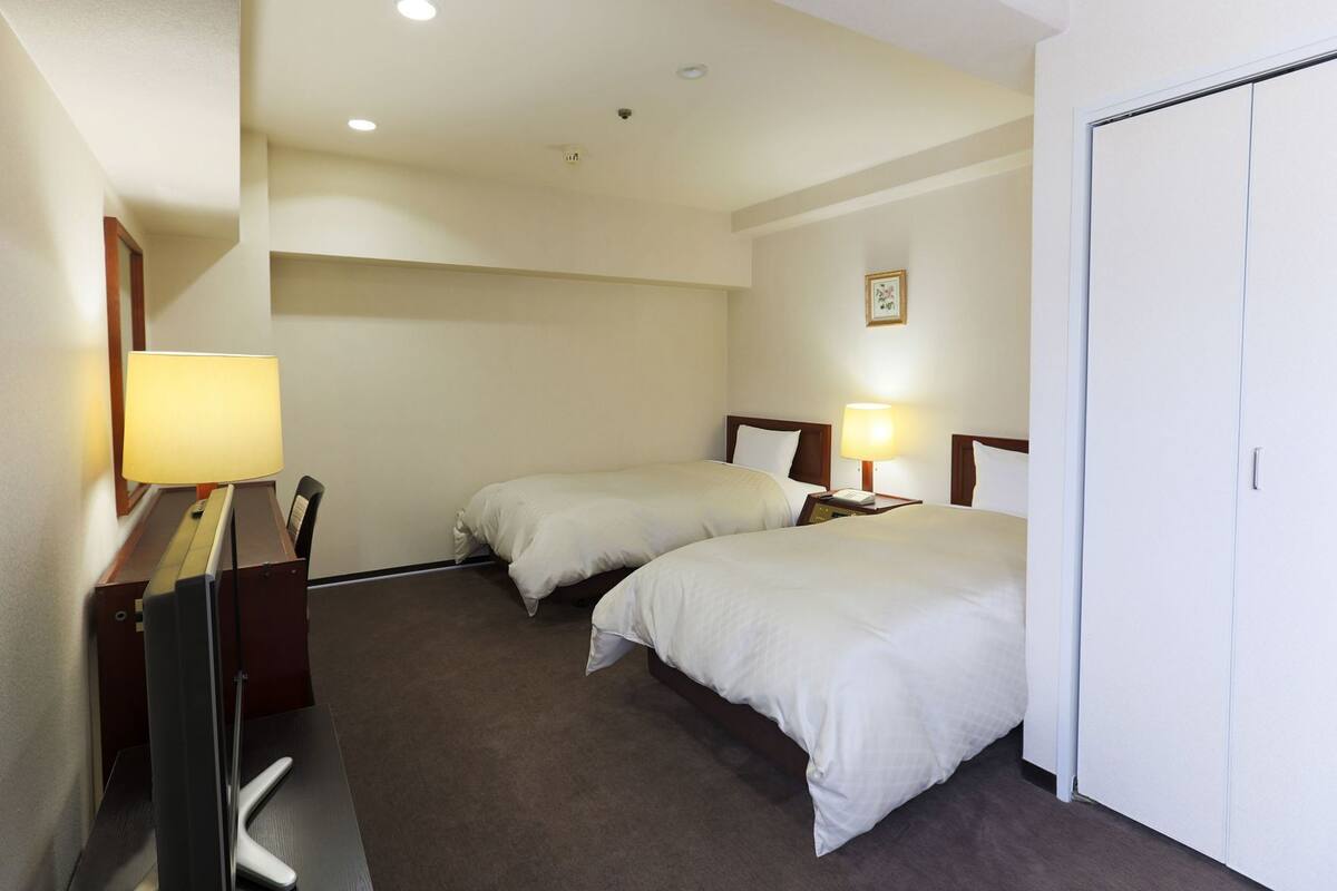 Quarto de hotel com duas camas, uma escrivaninha com abajur, um guarda-roupa e um quadro na parede.