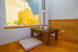 Um quarto com piso de madeira, uma mesinha com xícara e pires, uma janela com vista para uma cerca de bambu e um travesseiro no chão.