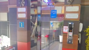 Um hall de hotel com balcão de recepção, porta de vidro de entrada, pilar vermelho e castanho, telefone e placa indicando área de serviço 24 horas.