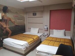 Quarto de hotel com duas camas, cada uma com colchão branco e manta dourada. Há uma prateleira fixada na parede com um telefone e uma pequena mesa ao lado das camas.