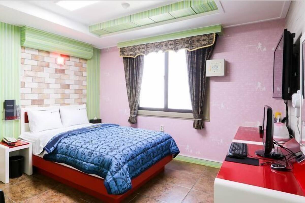 Quarto com parede em tijolos, cama com cobertor azul, escrivaninha com computador e janela com cortinas.