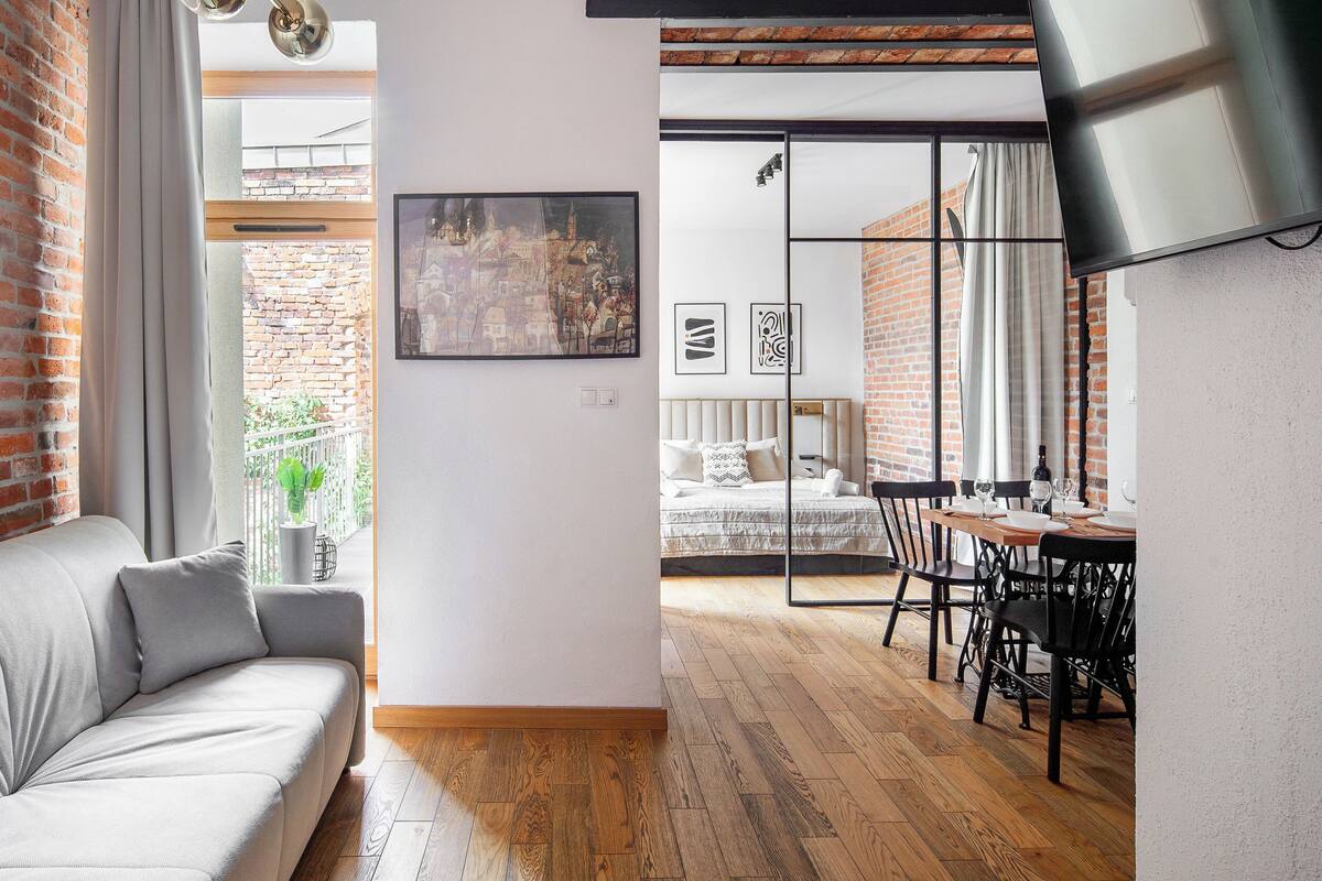 Uma sala de estar moderna com uma parede de tijolos, um sofá cinzento e uma área de jantar com mesa e cadeiras de madeira.