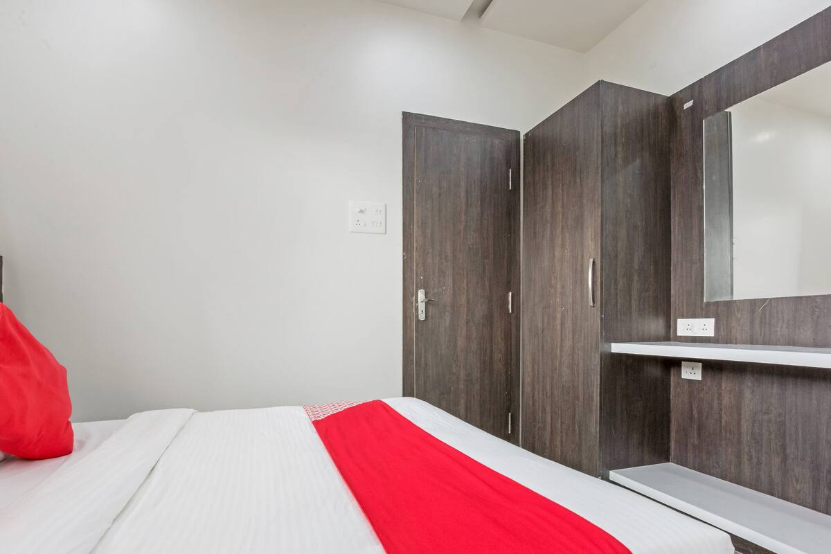 Quarto com cama, colcha vermelha e branca, guarda-roupa, espelho e prateleira.