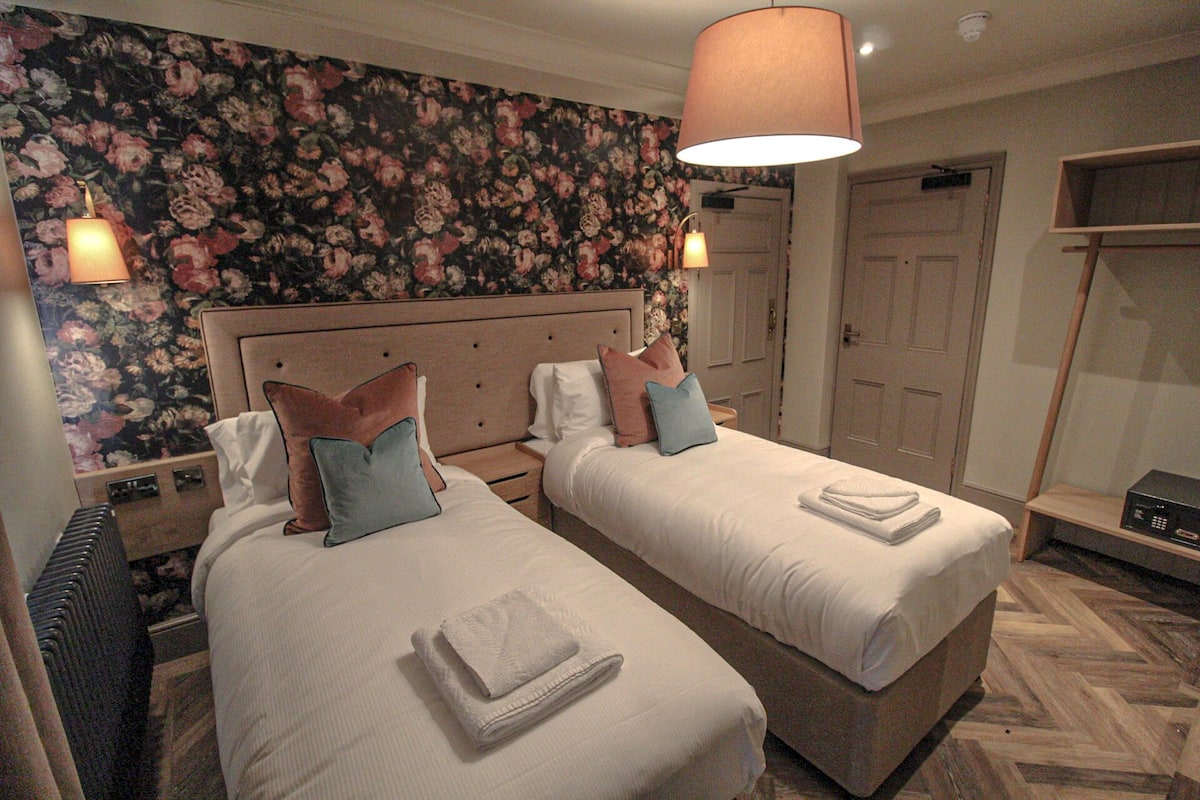 Quarto de hotel com duas camas, papel de parede floral, cabeceira de madeira, luminárias de apoio, guarda-roupa e televisão de tela plana.