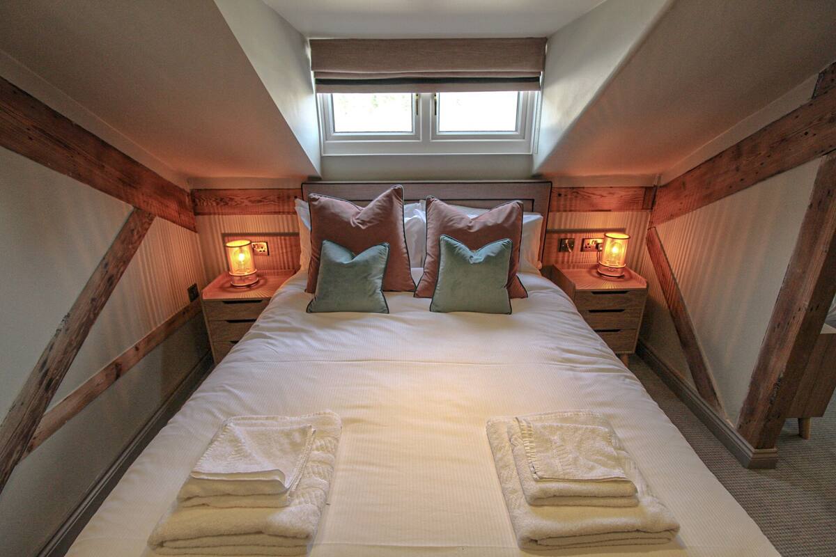 Quarto com uma cama grande, duas mesas de cabeceira com luminárias, uma janela com persianas e teto de madeira.