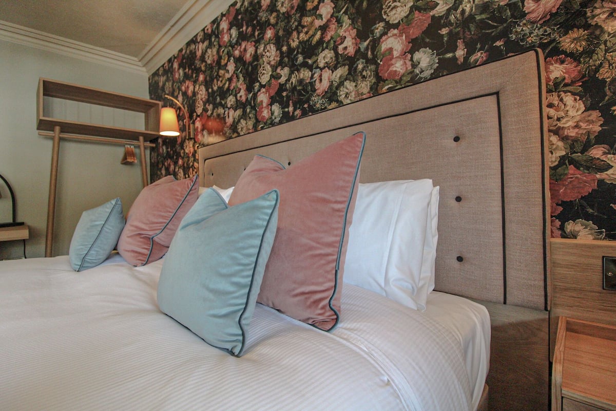 Cama com várias almofadas, cabeceira de madeira e papel de parede floral.