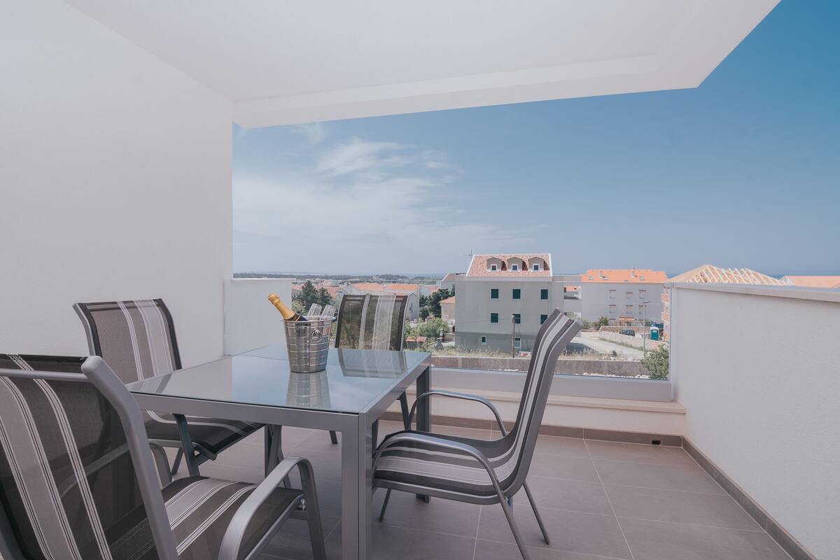 Uma varanda com mesa e cadeiras de vidro, vista para prédios e céu claro.
