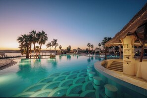 Piscina com vista para o pôr do sol, palmeiras e uma estrutura com telhado de palha.