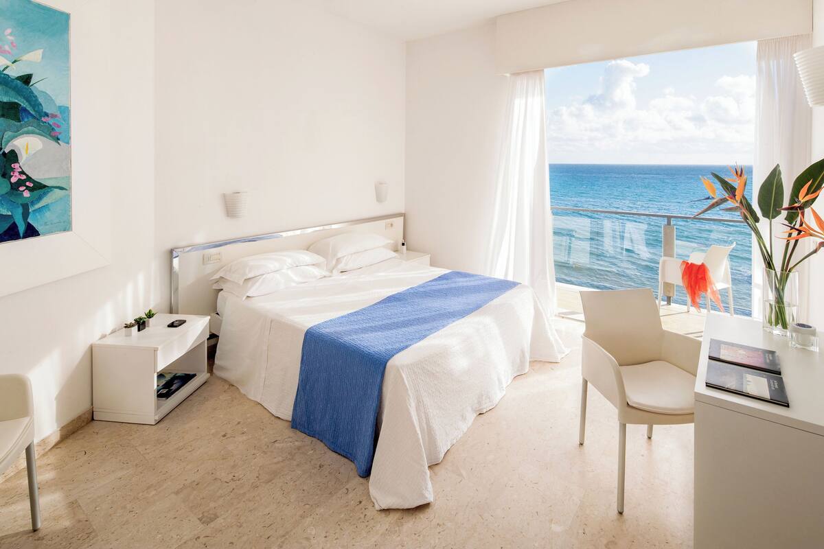 Quarto de hotel com cama, escrivaninha, cadeira e vista para o mar.