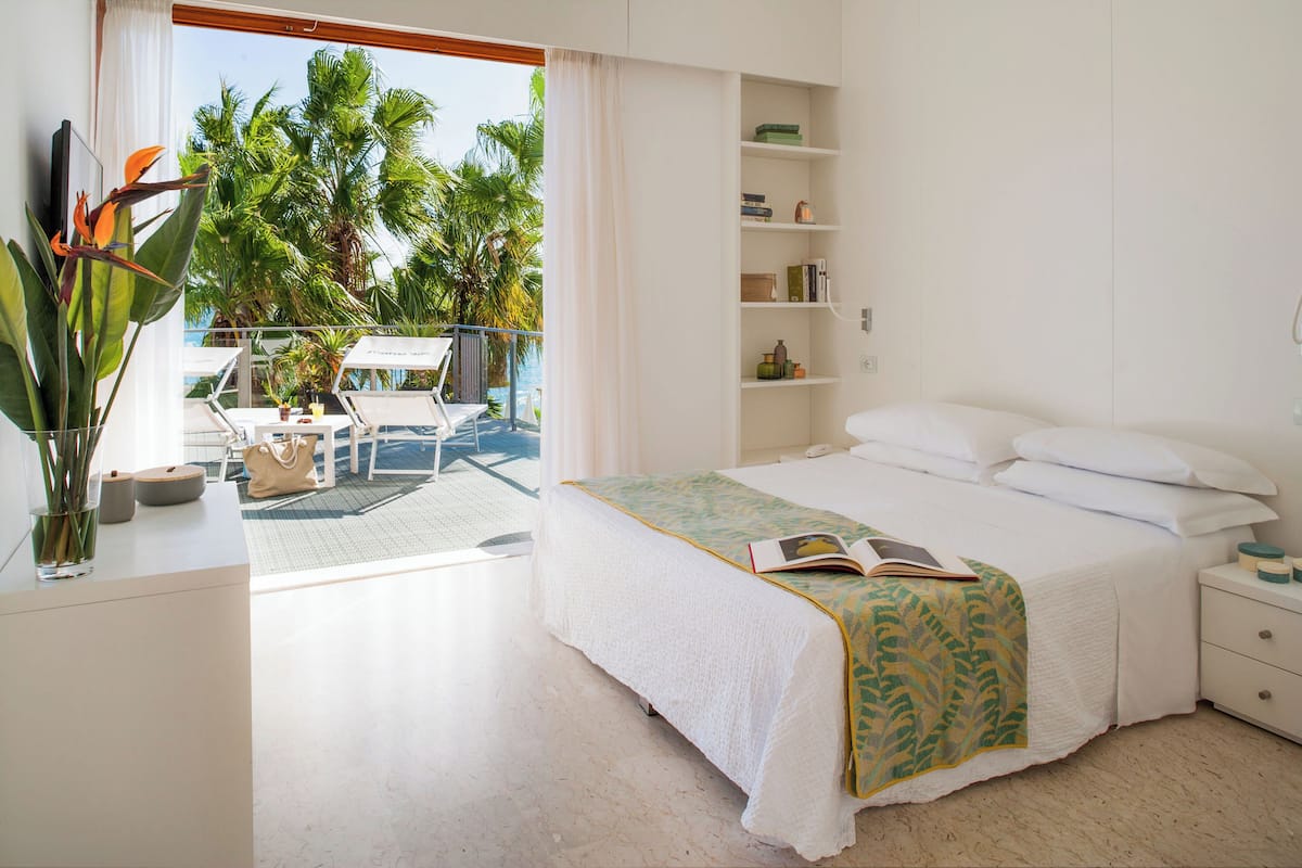 Quarto de hotel com cama, mesas de cabeceira, estante e vista para palmeiras e piscina.