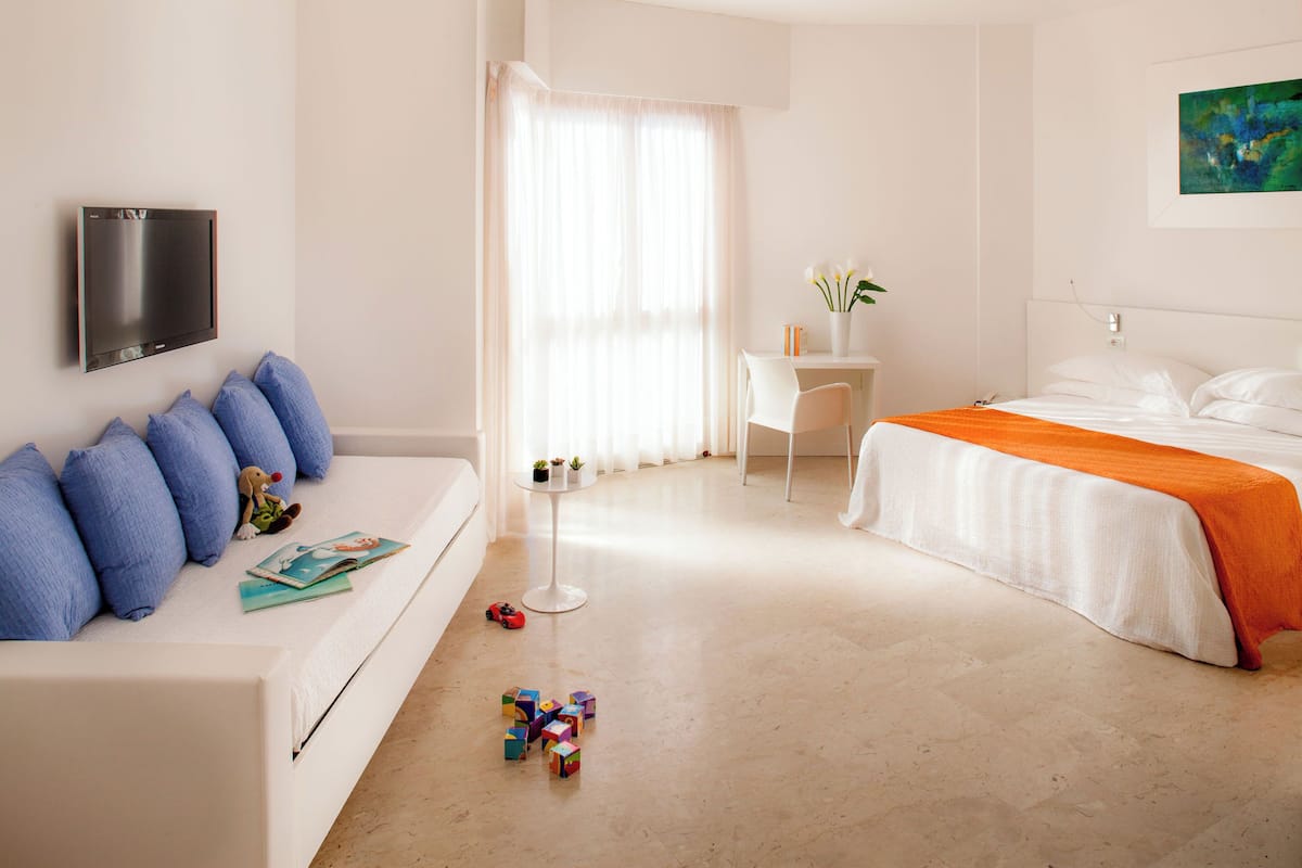 Quarto de hotel com uma cama branca, um sofá branco, uma pequena escrivaninha e uma televisão na parede.