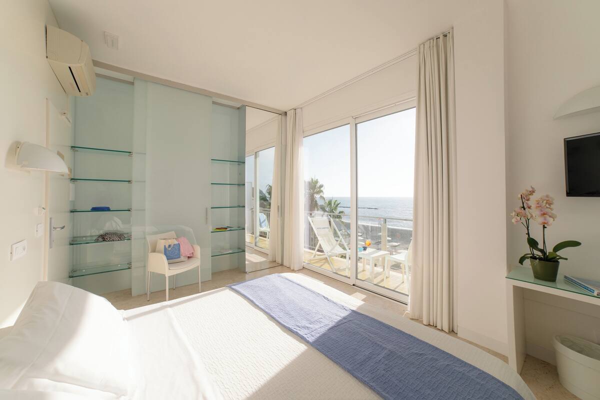 Quarto de hotel com uma cama grande, varanda envidraçada com vista para o mar, uma cadeira e televisão na parede.