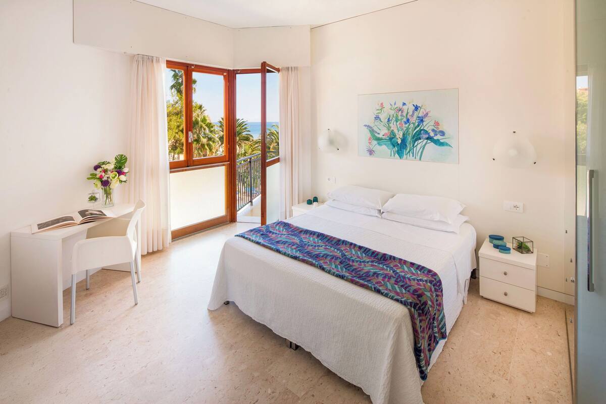 Quarto com cama, escrivaninha e varanda com vista para o mar.