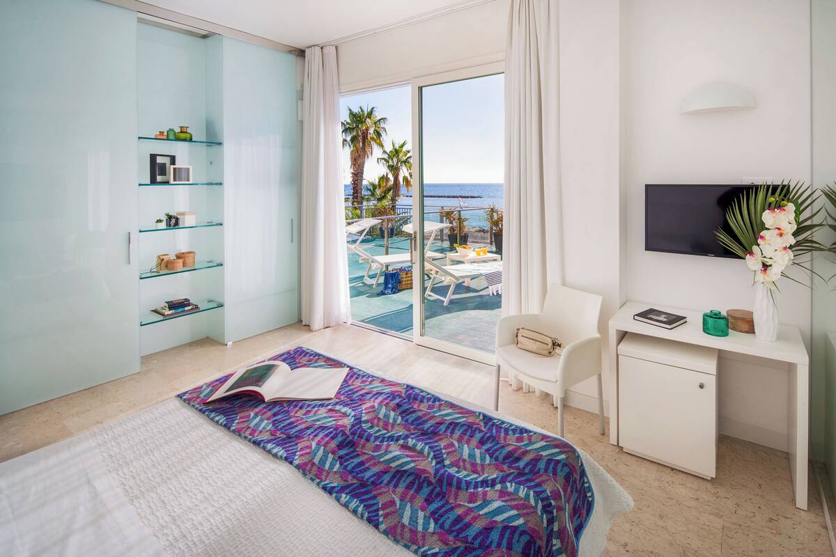 Quarto de hotel com cama, escrivaninha, televisão, cadeira, estante de livros e vista para o mar.