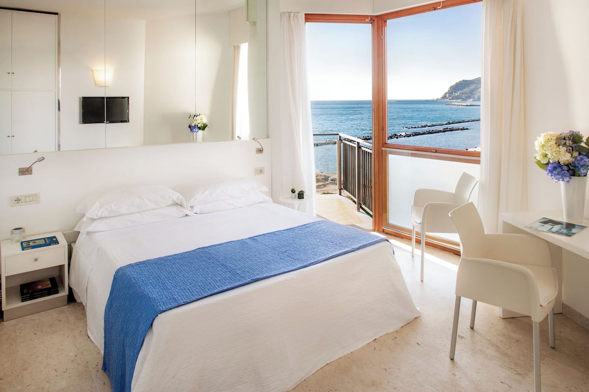 Quarto de hotel com uma cama grande, uma escrivaninha com cadeira, uma televisão e uma varanda vista para o mar.