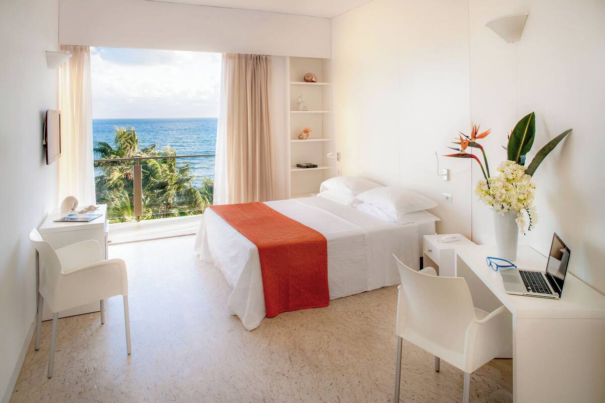 Quarto de hotel com cama, secretária com laptop, cadeira, prateleira pequena com objetos decorativos e vista para o oceano.