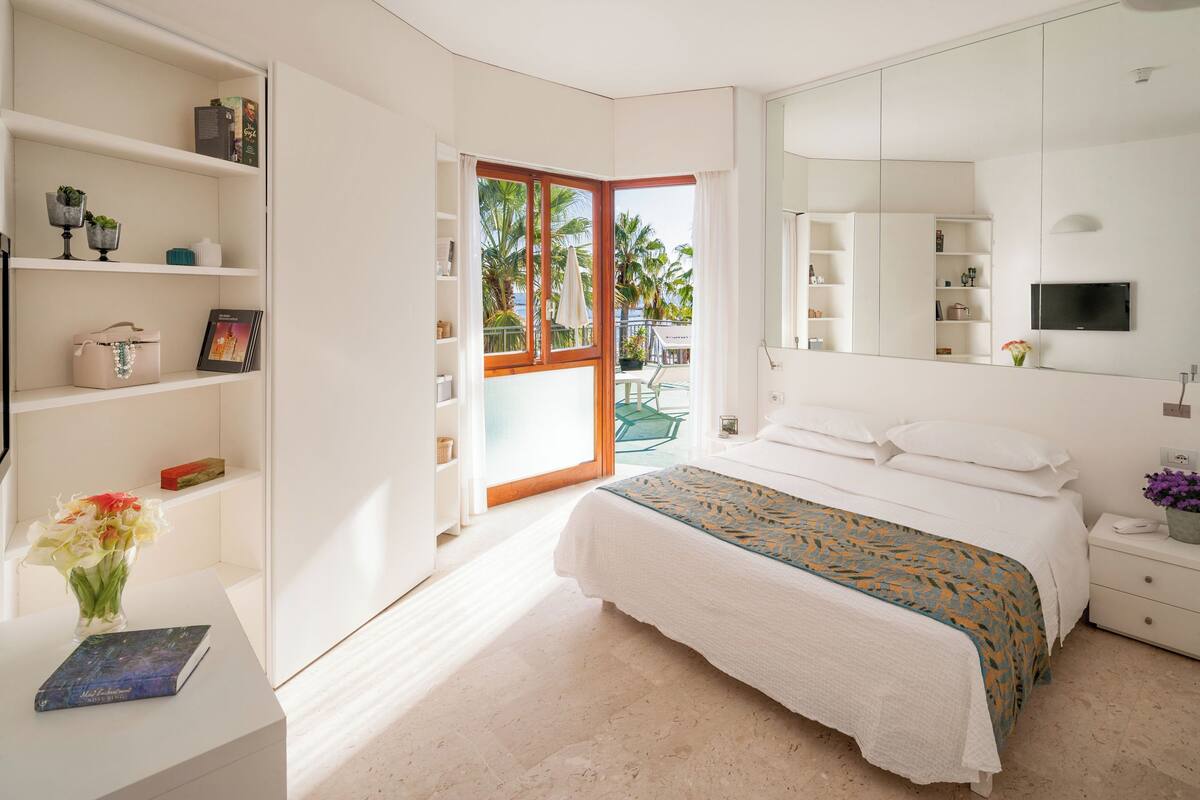 Quarto moderno com uma cama grande, um guarda-roupa branco, uma escrivaninha com um vaso de flores e vista para palmeiras por uma porta aberta.