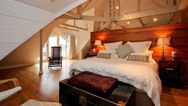 Um quarto com teto inclinado, vigas de madeira, uma cama grande com roupa de cama branca, duas mesas de cabeceira com abajures e uma cabeceira de madeira.