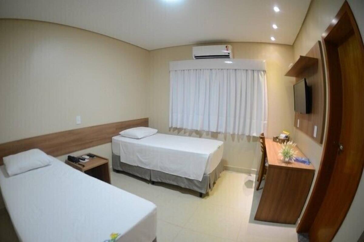 Quarto de hotel com duas camas de solteiro, uma escrivaninha com cadeira, televisão fixada na parede e uma janela com cortinas.