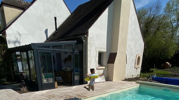 Piscina com água cristalina, rodeada por um deck de pedra e uma casa moderna com grandes janelas e um alpendre coberto.
