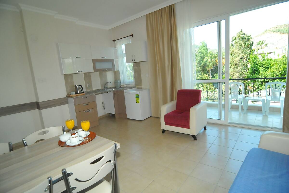 Quarto de hotel moderno com área de refeições, cozinha compacta e varanda com vista para áreas verdes.
