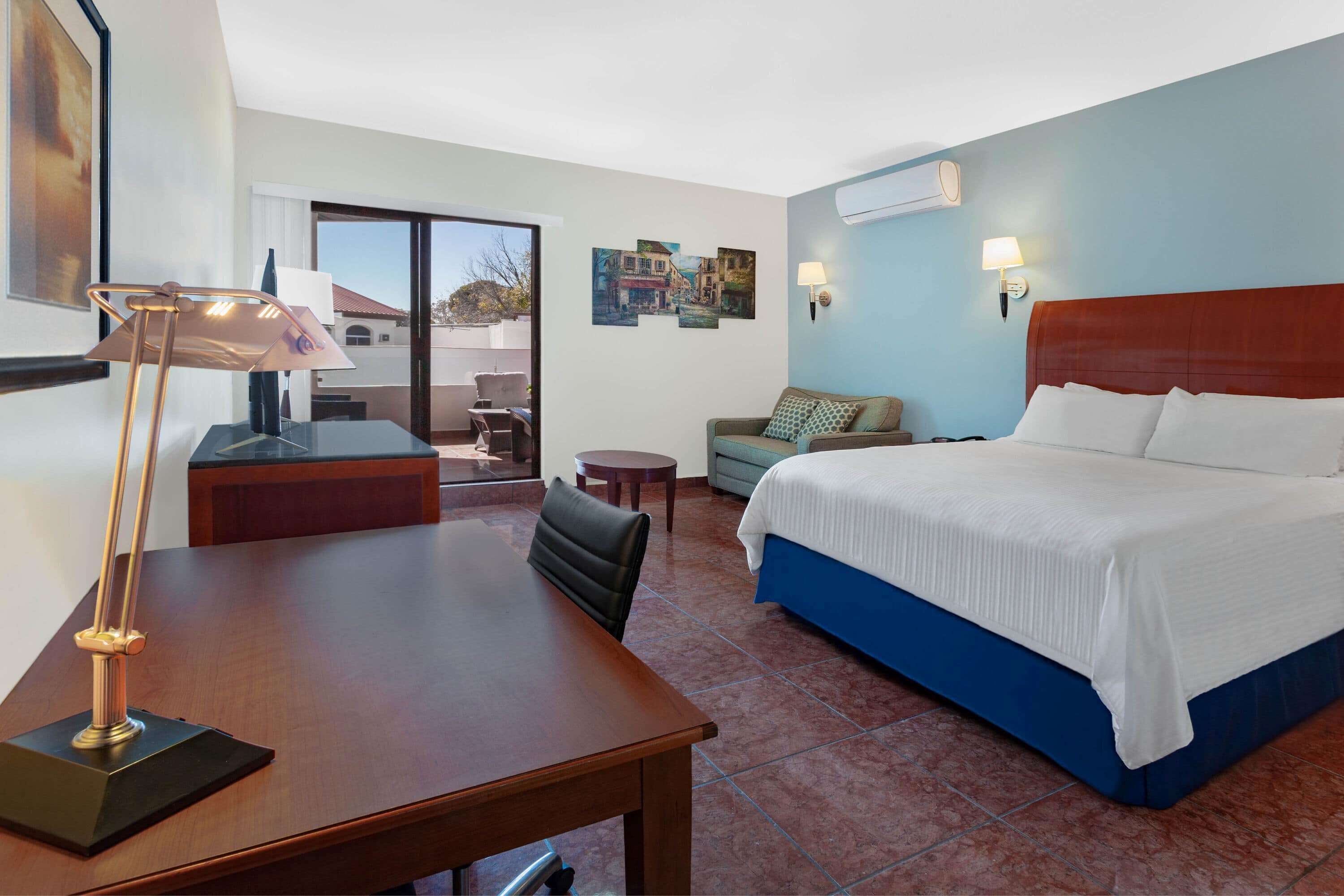 Quarto de hotel com cama, escrivaninha, cadeira, mesinha, sofá e varanda com vista.