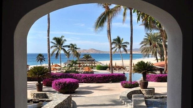 Visão através de um arco de um resort, mostrando uma piscina, palmeiras e uma praia.