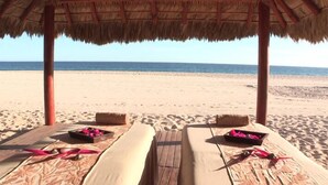 Um espaço de relaxamento à beira-mar, com bancos de madeira e telhado de palha, com vista para uma praia arenosa e o oceano.