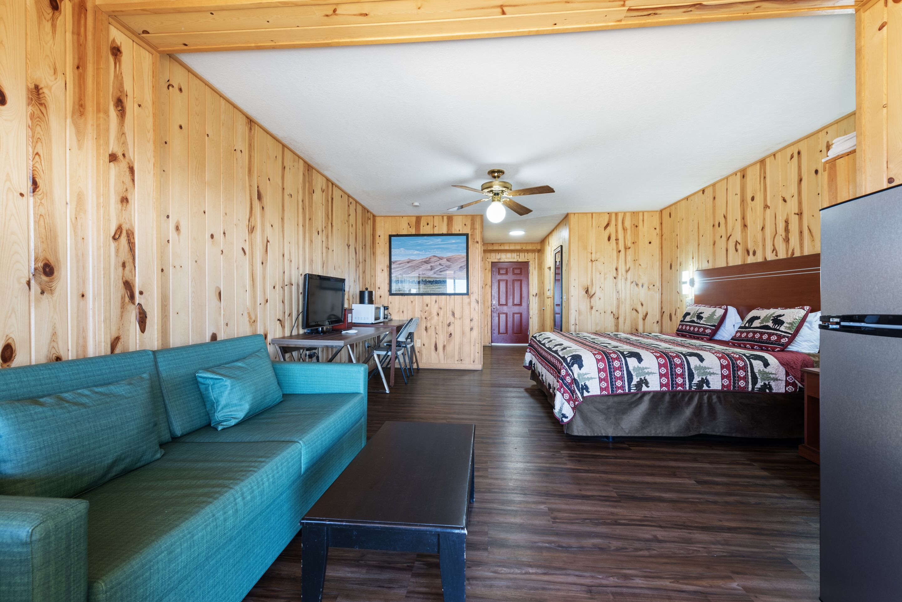 Quarto com decoração em madeira, uma cama com roupa de cama estampada, um sofá, uma escrivaninha com um computador e um ventilador de teto.
