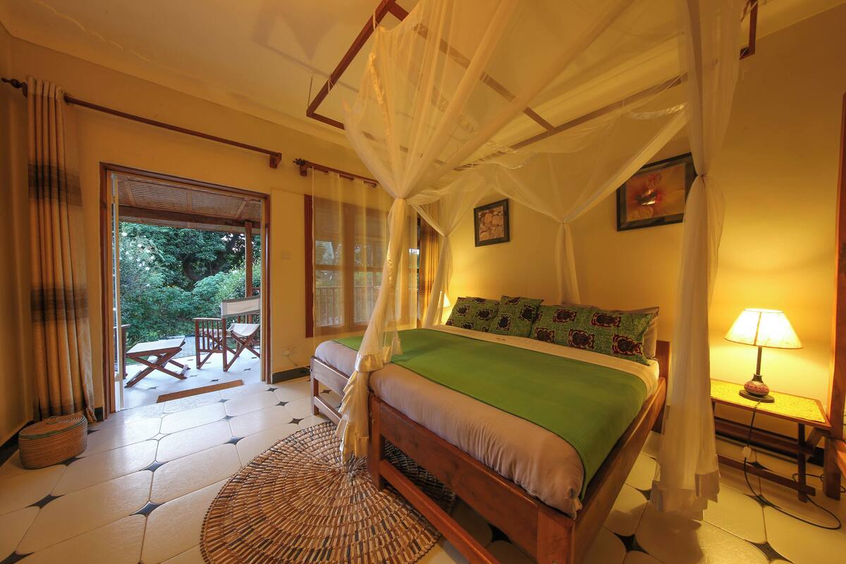 Quarto com cama, mesa de cabeceira com abajur, janela com cortinas e vista para o exterior.