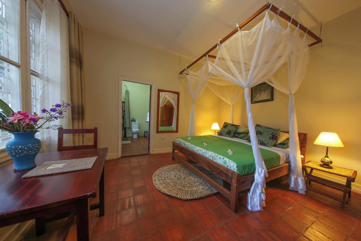 Quarto com cama de dossel, mesa de cabeceira com abajur, cadeira de madeira, tapete redondo pequeno e um vaso com flores sobre uma mesa de madeira.