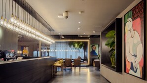 Uma área de recepção moderna com um balcão comprido, banquetas e uma máquina de café. Há luminárias no teto e uma planta grande ao fundo.