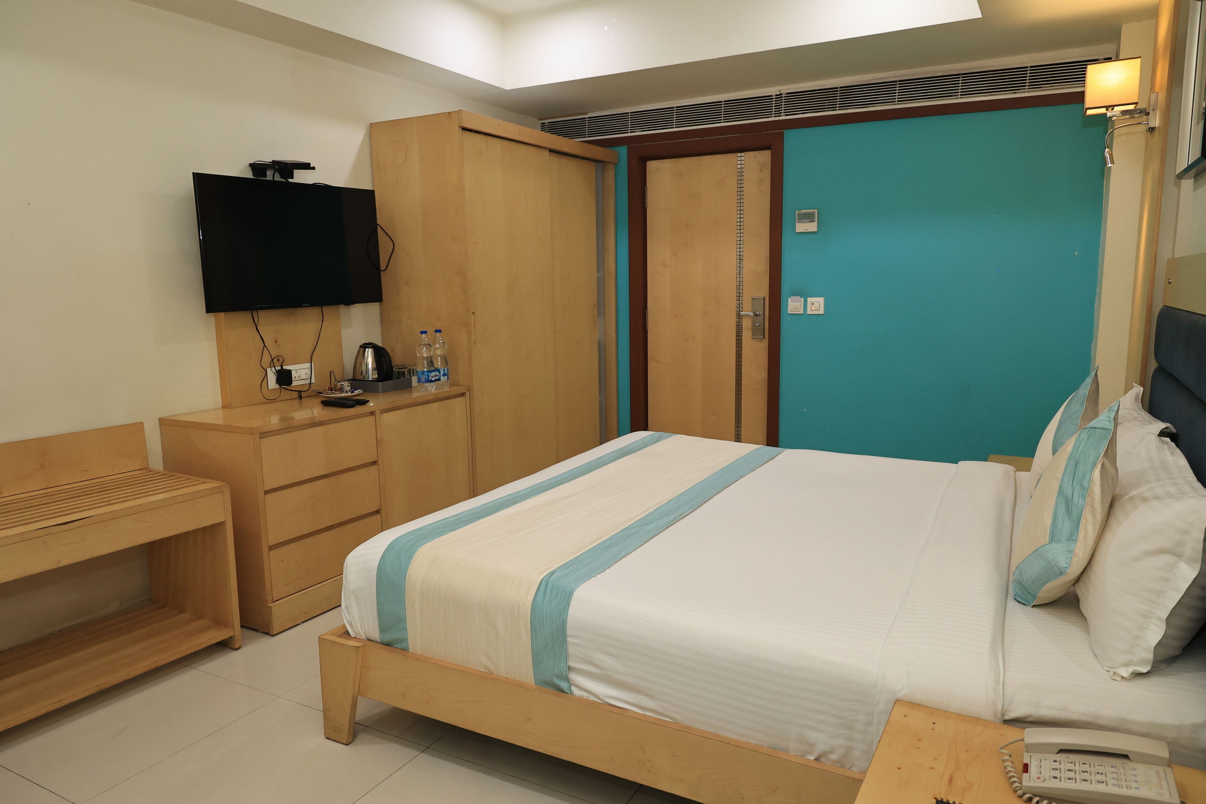 Quarto de hotel com cama, escrivaninha de madeira, televisão, guarda-roupa e porta de banheiro.