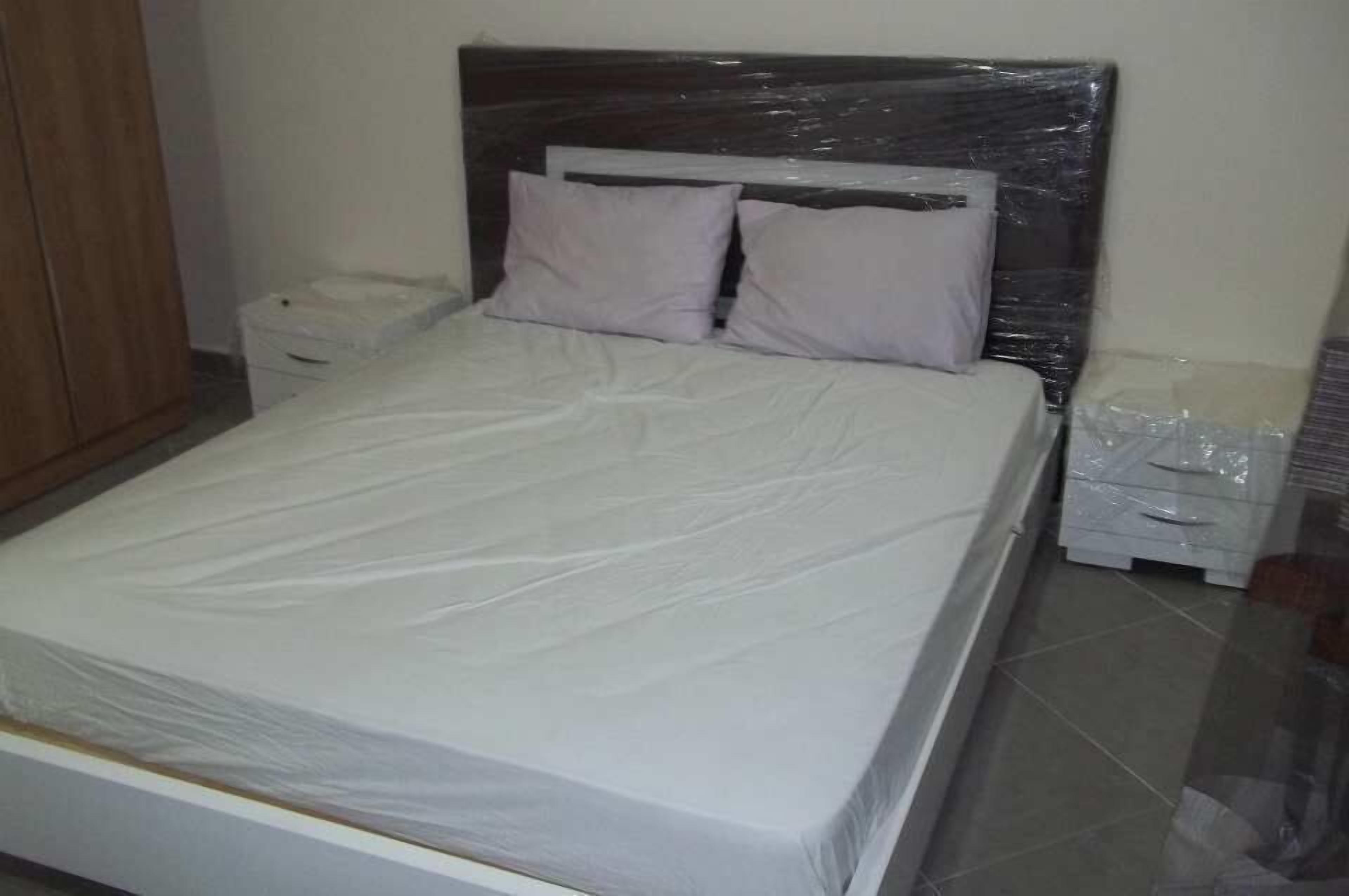 Uma cama bem arrumada com colchão e travesseiros brancos, cabeceira de madeira e mesa de cabeceira branca com abajur.