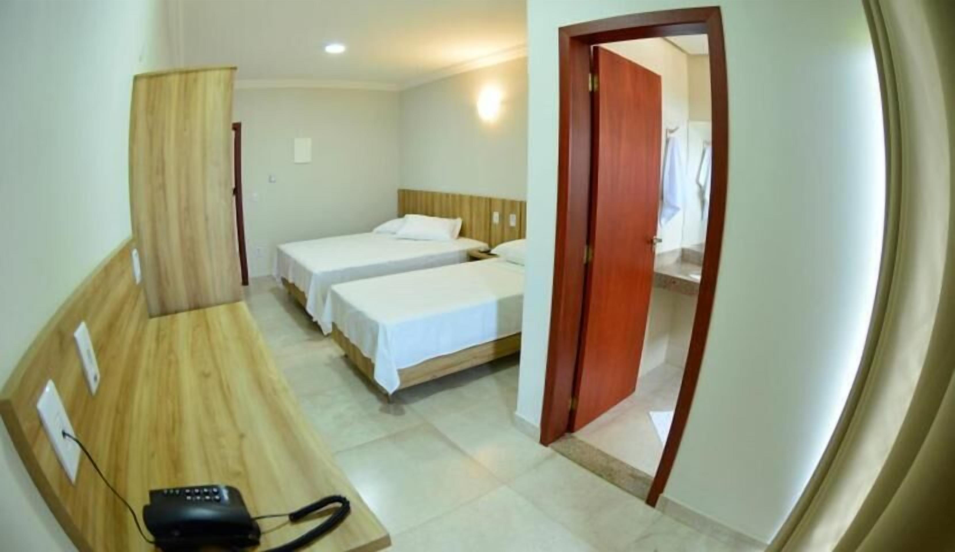 Quarto com duas camas de solteiro, uma escrivaninha de madeira, uma cadeira e uma mesinha com controle remoto.