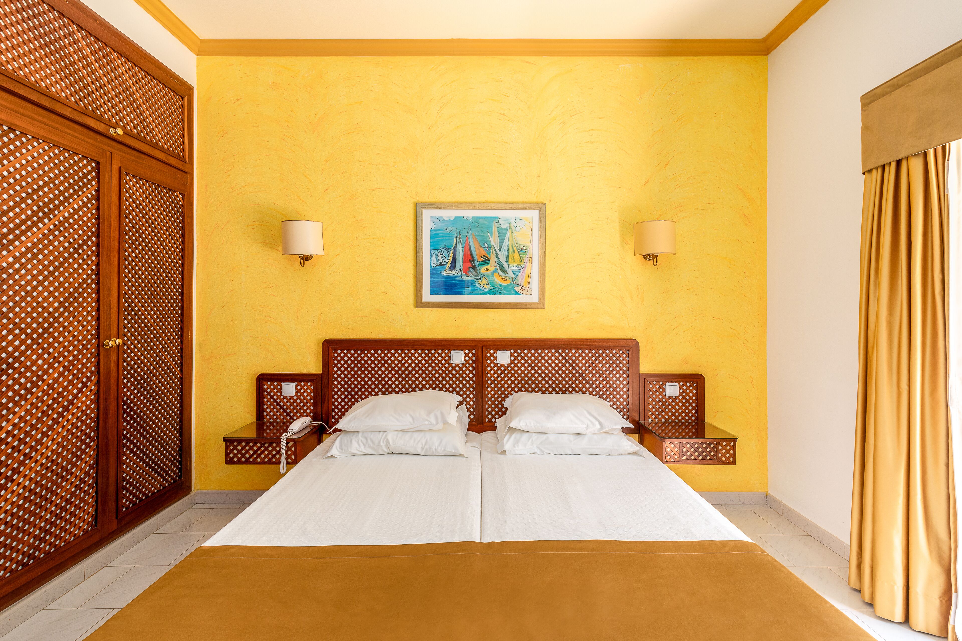 Quarto de hotel com cama de casal, cabeceira de madeira, duas mesas de cabeceira com abajures, um quadro na parede e uma cortina na janela.