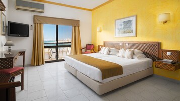 Quarto de hotel com uma cama grande, televisão, escrivaninha com luminária, cadeira e varanda com vista para o mar.