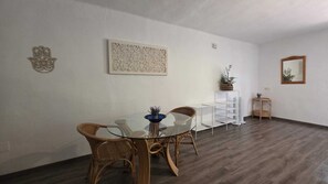 Um quarto com uma mesa de vidro cercada por cadeiras de vime, uma estante branca, uma planta em vaso e um espelho na parede.