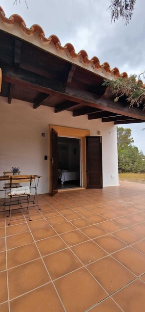 Uma varanda coberta com piso de azulejos, uma mesa de madeira com cadeiras e uma porta que dá para o interior.