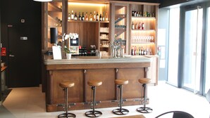 Uma área de bar com banquetas de madeira, prateleiras com garrafas e copos.