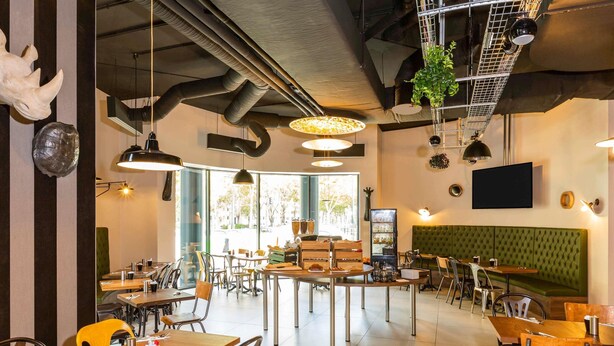 Um restaurante com várias mesas, cadeiras, um banco estofado verde e iluminação pendente, além de uma grande janela que oferece vista para o exterior.